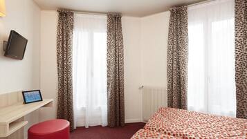 Quarto de hotel com cama, um banquinho vermelho, televisão em uma prateleira e cortinas de onça.