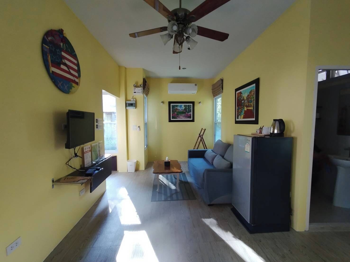 Sala de estar com televisão, sofá e frigorífico, com paredes amarelas e ventoinha no teto.