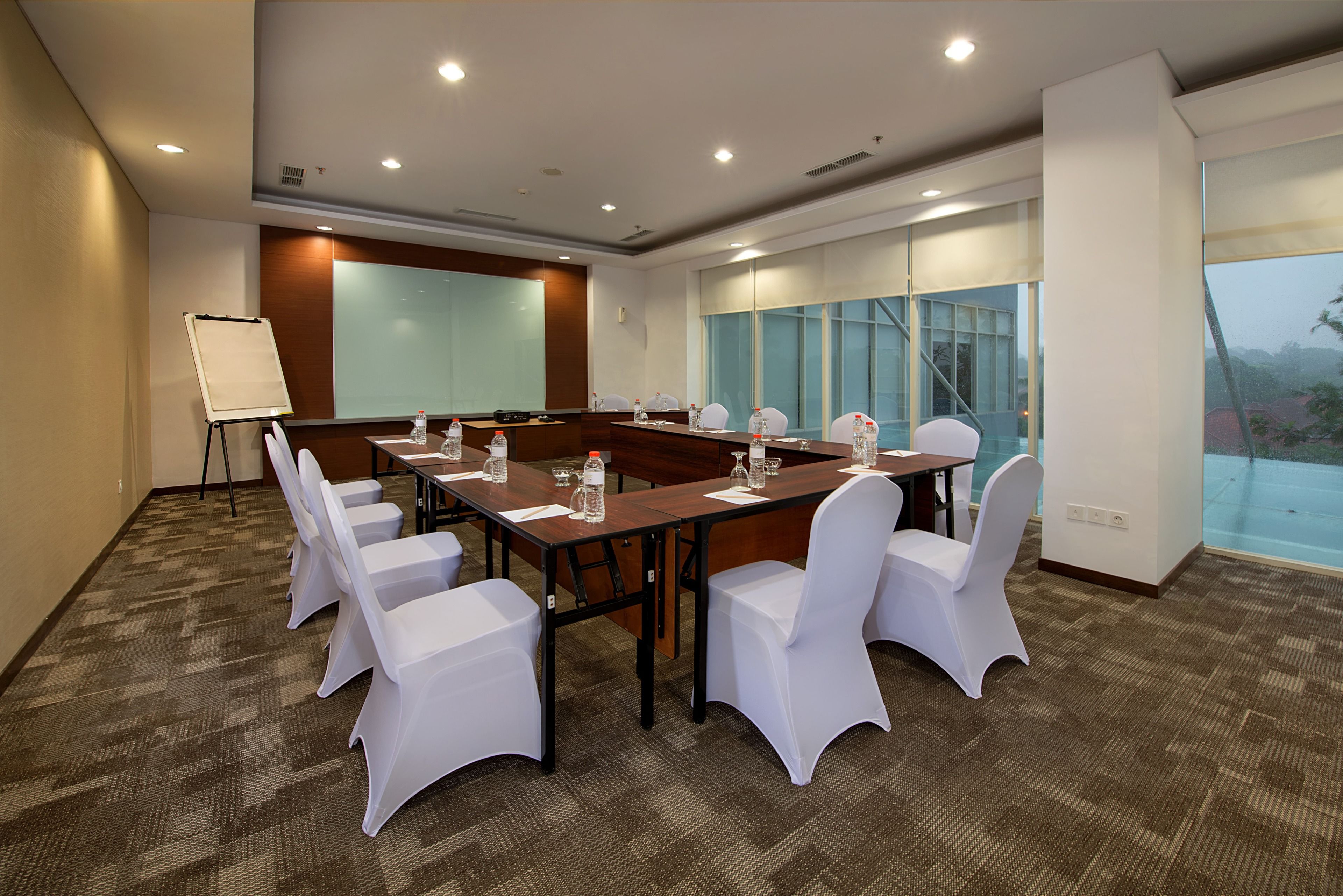 Uma sala de conferência com uma mesa grande, cadeiras brancas, um quadro branco e vista para o exterior através de grandes janelas.