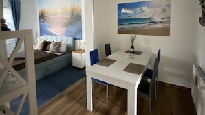 Quarto com mesa de jantar, cadeiras, cama e vista para a praia, e uma pintura do oceano.