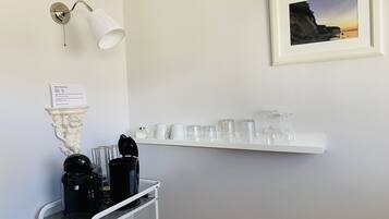 Quarto de hotel com parede branca, máquina de café embutida, prateleira branca com copos e quadro com paisagem costeira.