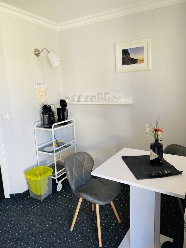 Quarto pequeno e limpo com uma mesa branca, uma cadeira cinza, uma máquina de café, uma estante com copos e um quadro na parede.