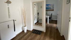 Um corredor com piso de madeira, um radiador branco, uma planta em vaso, um espelho e uma porta que dá para um quarto com uma pintura.