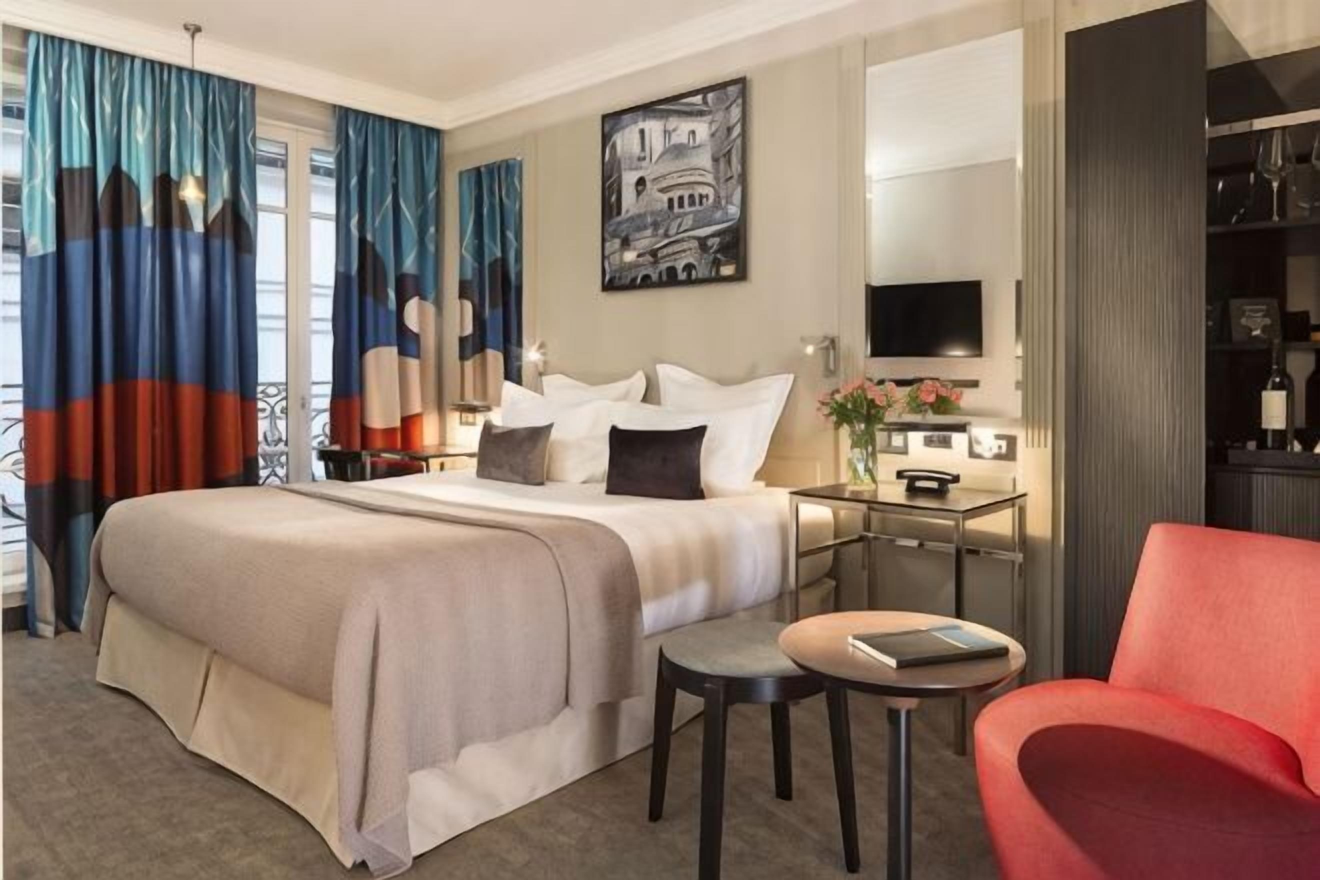 Quarto de hotel com cama, escrivaninha, cadeira, televisão e um quadro na parede.