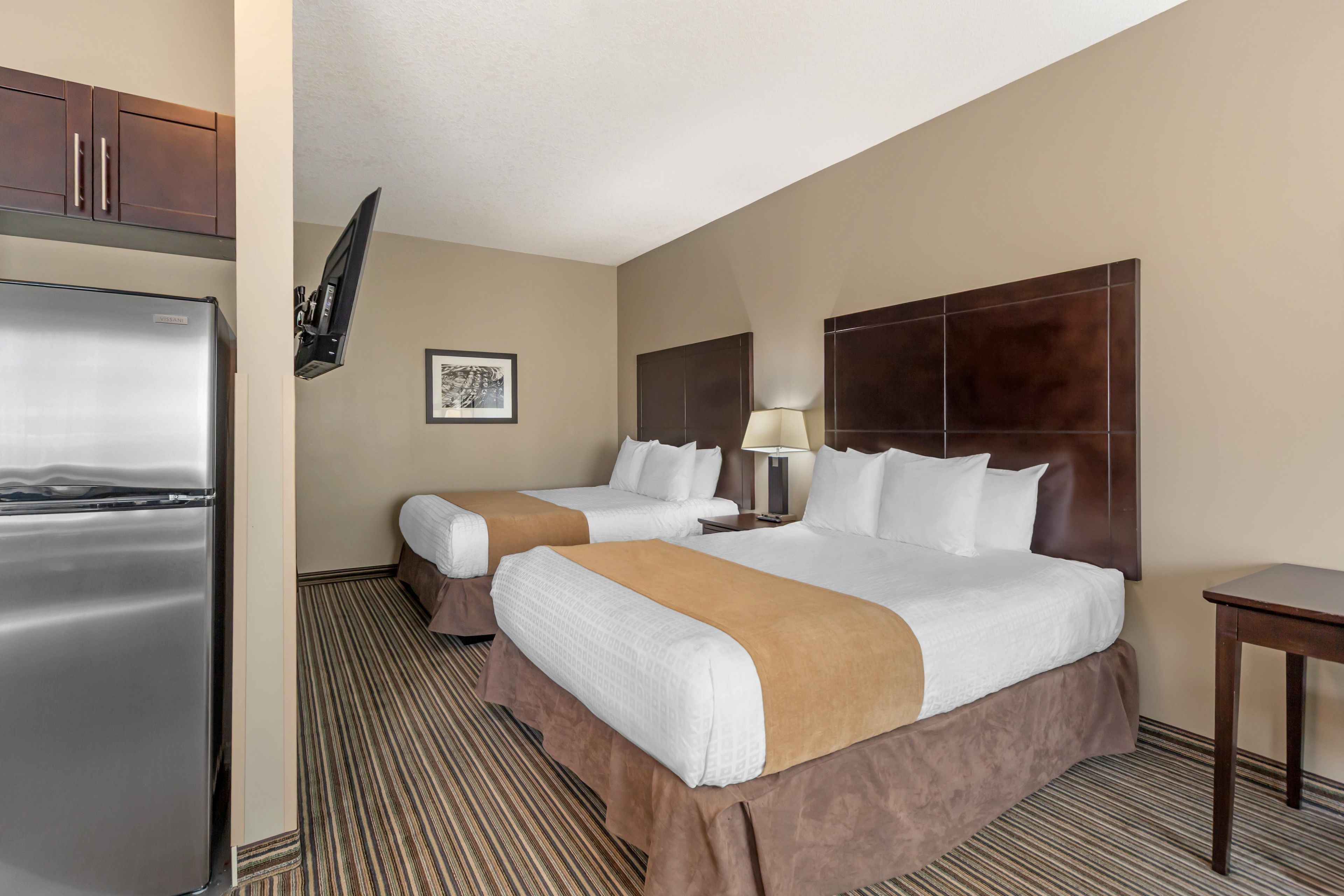 Quarto de hotel com duas camas, um frigorífico, uma mesa de cabeceira com uma lâmpada e um quadro pendurado na parede.