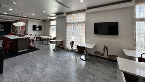 Um hall de hotel moderno com área de bar central, diversos arranjos de assentos e uma grande tela de televisão.