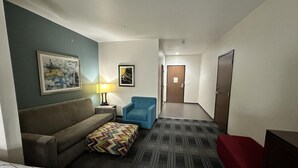Quarto de hotel com sofá, poltrona azul, banquinho estampado, luminária e uma porta ao fundo.
