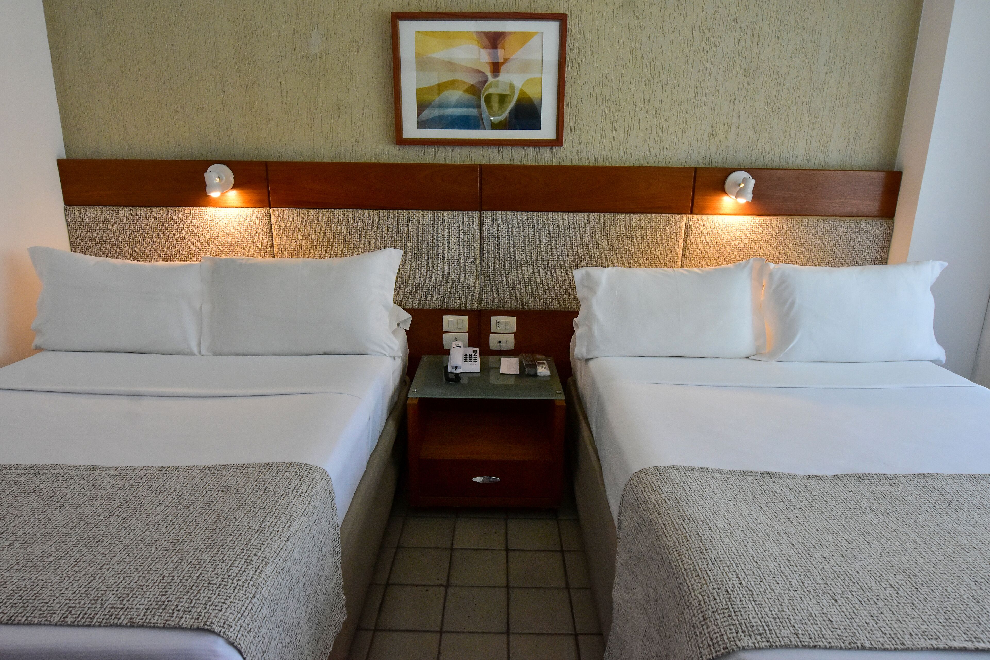Quarto de hotel com duas camas de solteiro, uma mesa de cabeceira com telefone e luminária, e um quadro na parede.