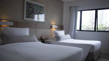 Quarto de hotel com duas camas, cabeceira com luminária, quadro na parede e janela com vista para edifícios.