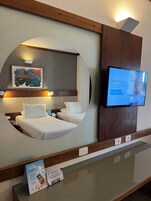 Quarto de hotel com uma cama, uma TV fixada na parede, uma obra de arte emoldurada acima da cama e uma receção com um aviso.