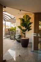 Um saguão moderno de hotel com uma grande planta em vaso, uma porta de vidro com um logotipo e uma parede revestida em azulejos.