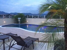 Piscina com borda revestida, cercada por um deck de madeira e cadeiras de metal.
