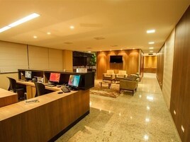 Uma área de recepção de hotel com um balcão de madeira, vários monitores de computador e uma área de estar com cadeiras e um sofá.