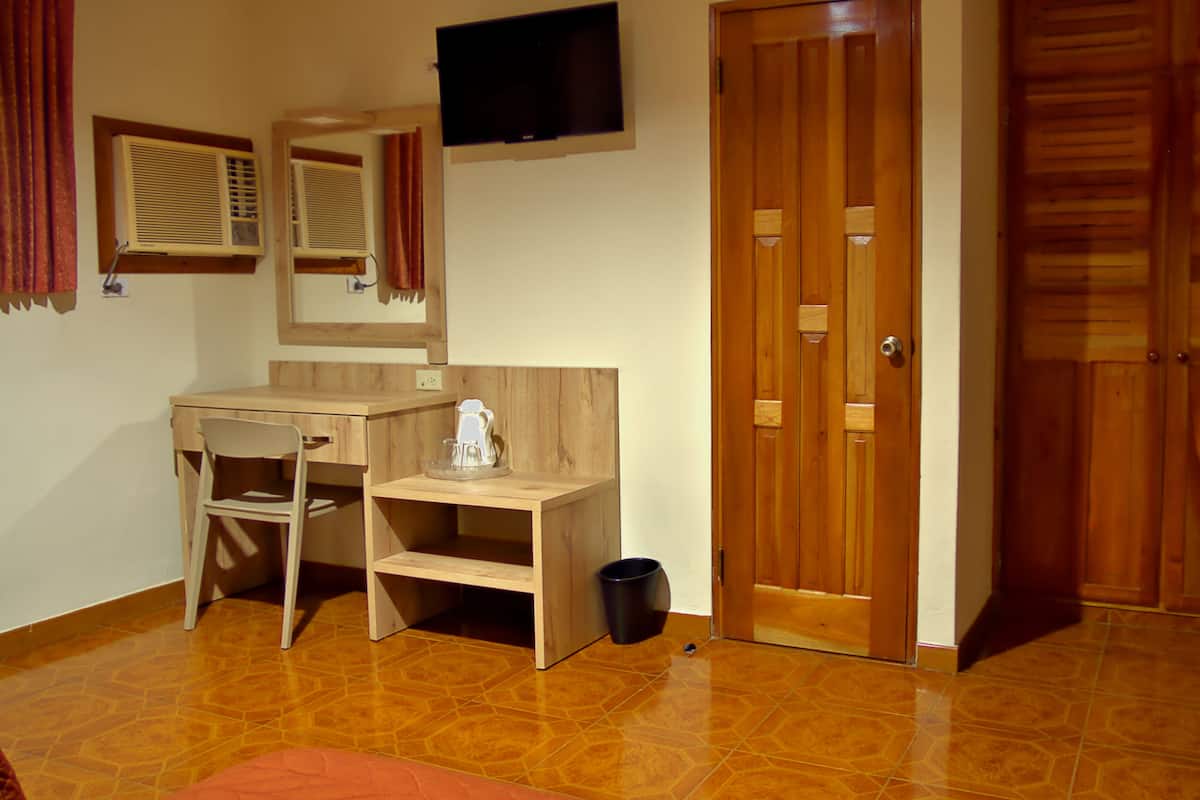 Quarto com escrivaninha de madeira, cadeira e televisão fixada na parede. Há um guarda-roupa de madeira com porta e uma lixeira ao lado.
