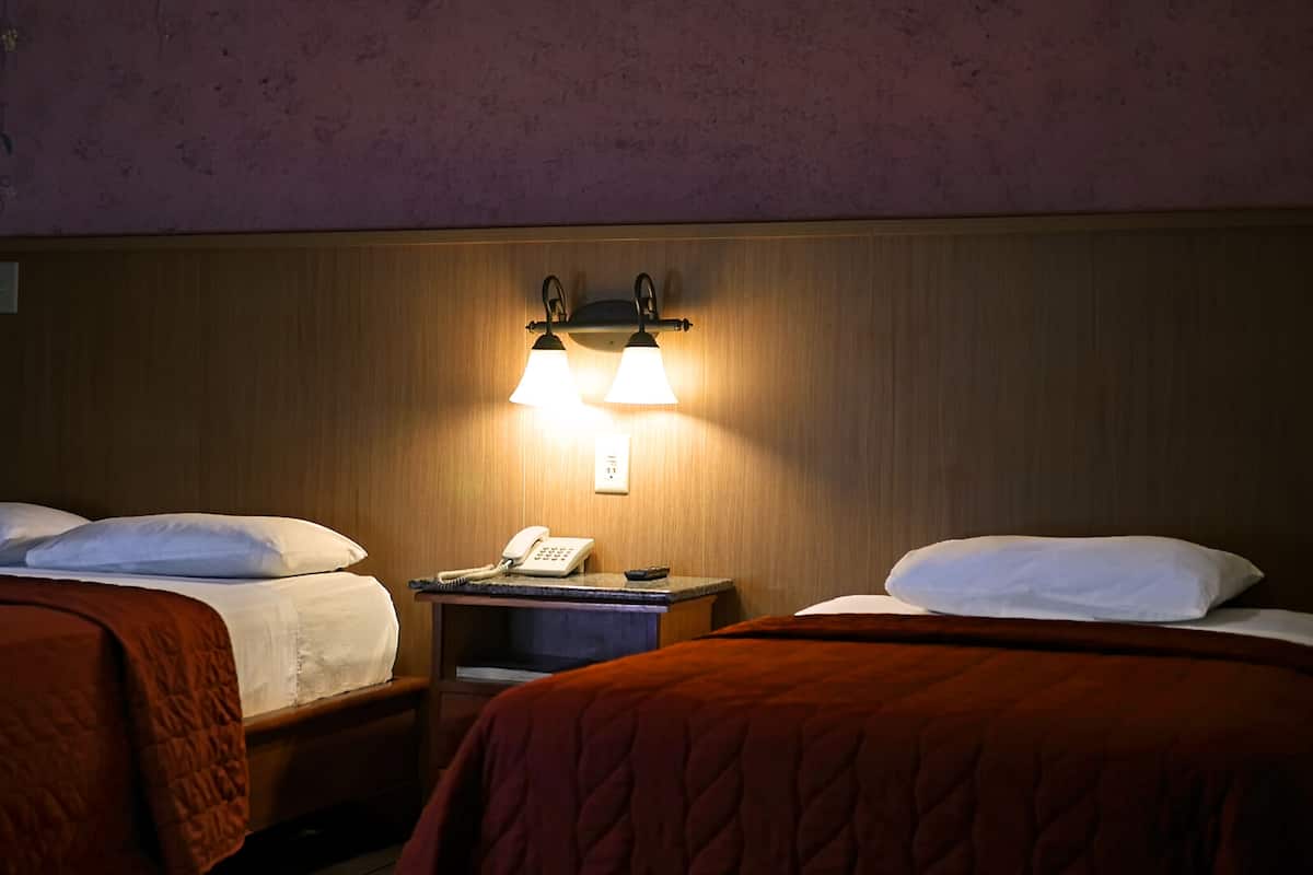 Quarto de hotel com duas camas, uma mesa de cabeceira com telefone e luminária na parede.