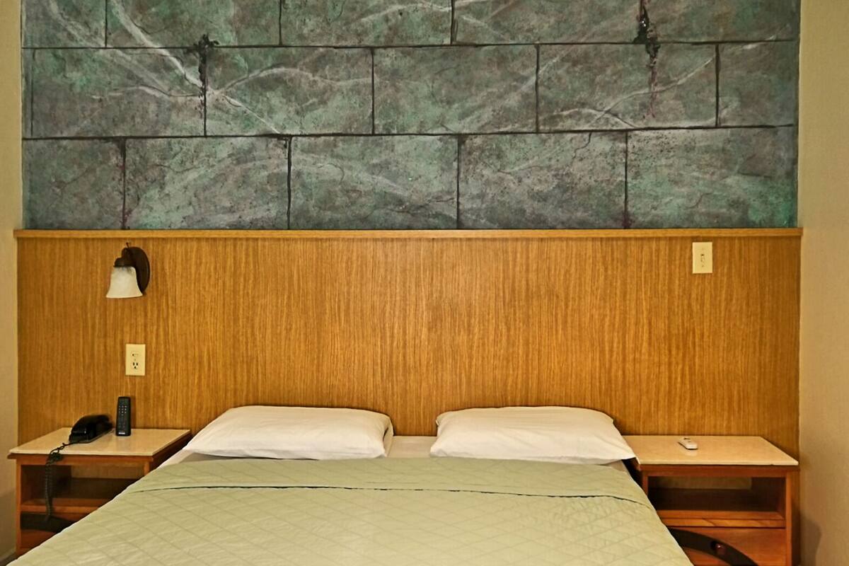 Quarto de hotel com cabeceira de madeira, duas mesas de cabeceira, cama com colcha verde, luminária na parede e telefone.