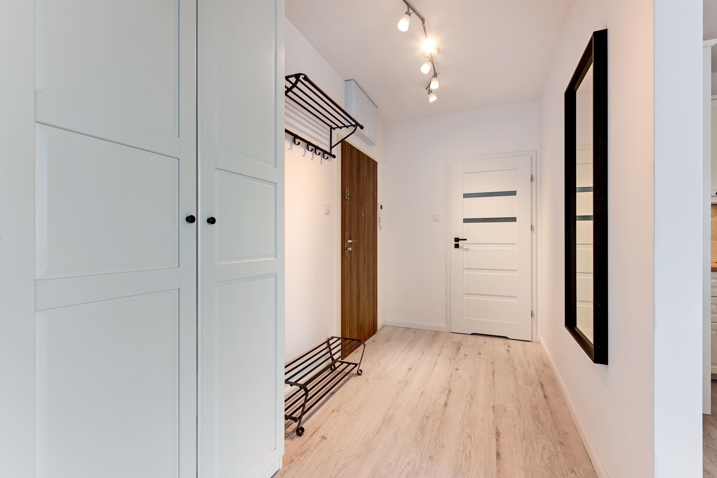 Um corredor com portas brancas, uma porta de madeira, uma estante de metal e um espelho fixado na parede.