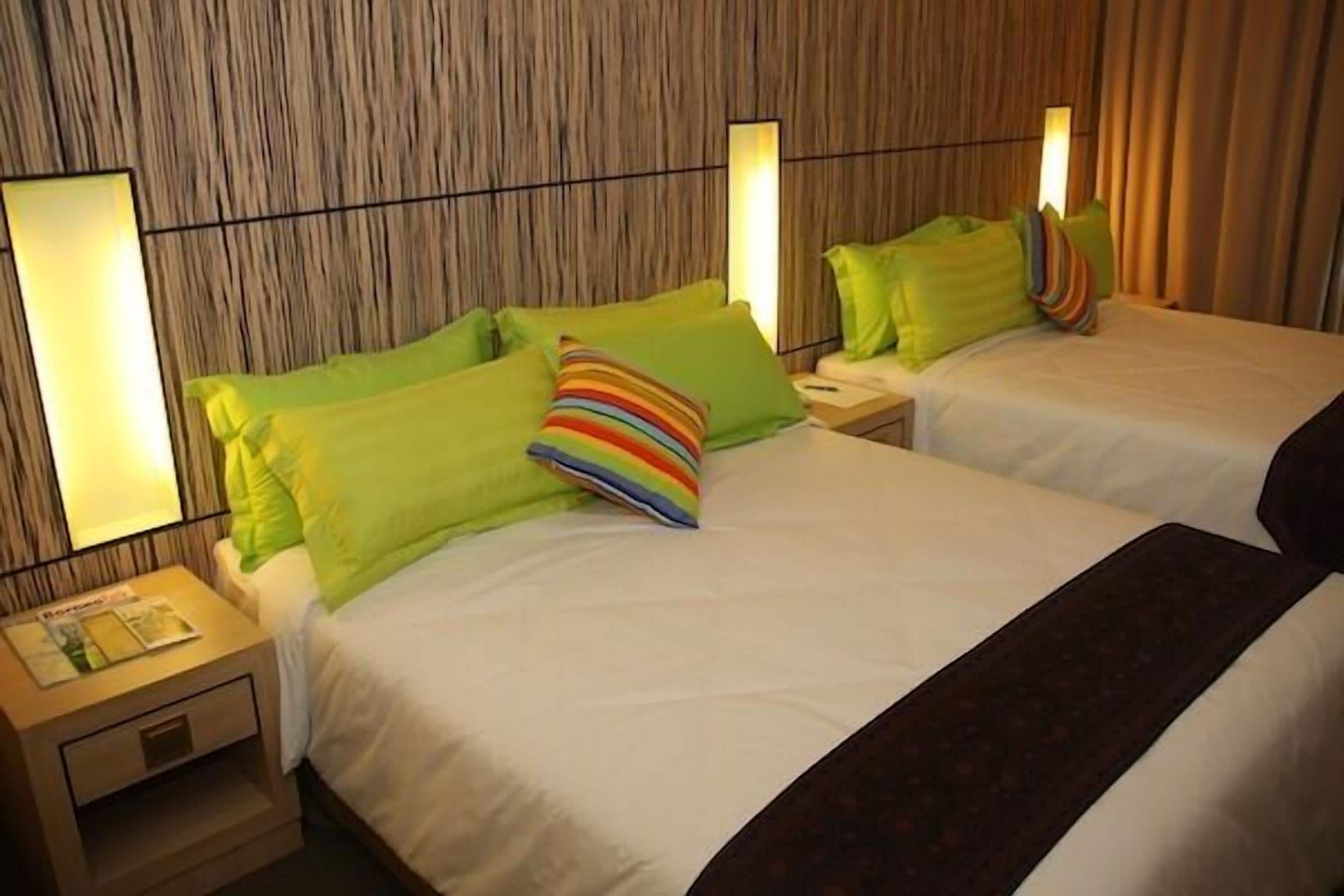 Quarto de hotel com duas camas, cada uma com almofadas verdes e almofadas listradas. Aparelhadores de cabeceira com abajures e uma revista em um deles.