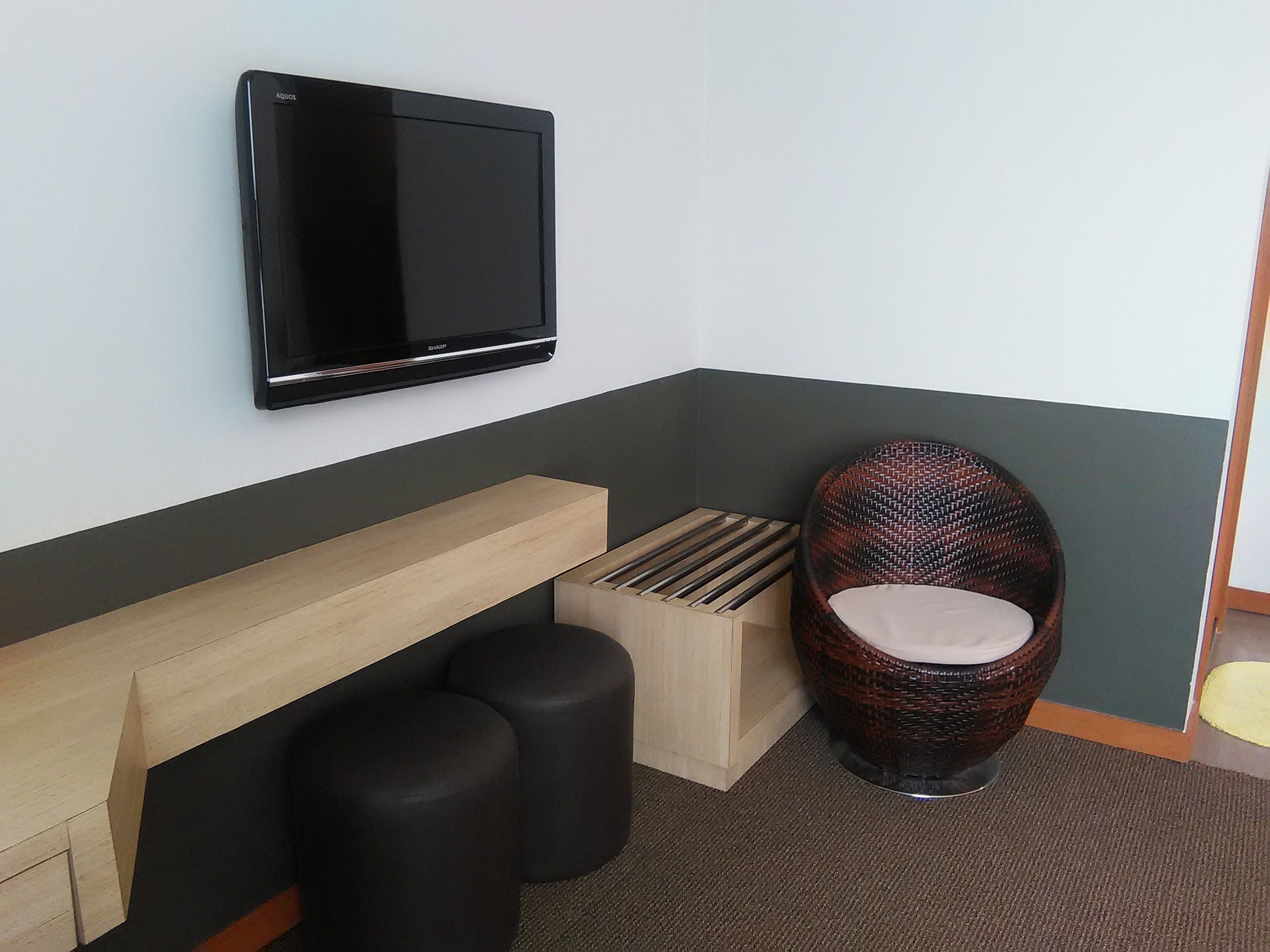 Quarto moderno com TV de tela plana na parede, um banco de madeira, dois pufes pretos e uma cadeira de vime com almofada.
