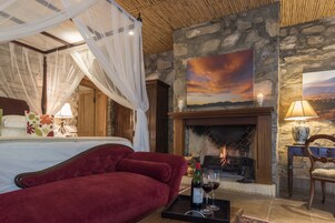 Quarto com sofá de veludo vermelho, cama com dossel, lareira de pedra e pintura de um pôr do sol.