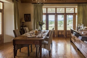 Uma sala de jantar com uma mesa de madeira posta para quatro pessoas, cercada por cadeiras. Há um vaso com flores sobre a mesa, além de garrafas de vinho e copos. O ambiente possui piso de madeira e uma vista para o exterior através de grandes portas de vidro.