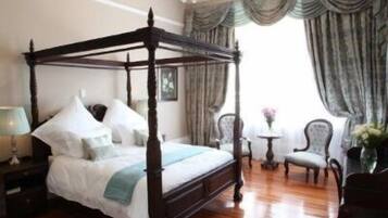 Uma cama de dossel com um candelabro, uma mesa redonda pequena e duas cadeiras num quarto com soalho de madeira e cortinas.