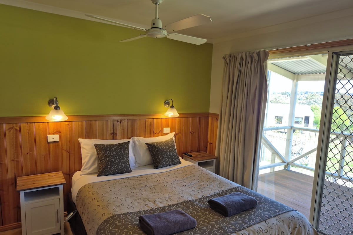 Quarto com cabeceira de madeira, duas luminárias laterais, um ventilador de teto e uma cama grande com roupa de cama estampada.