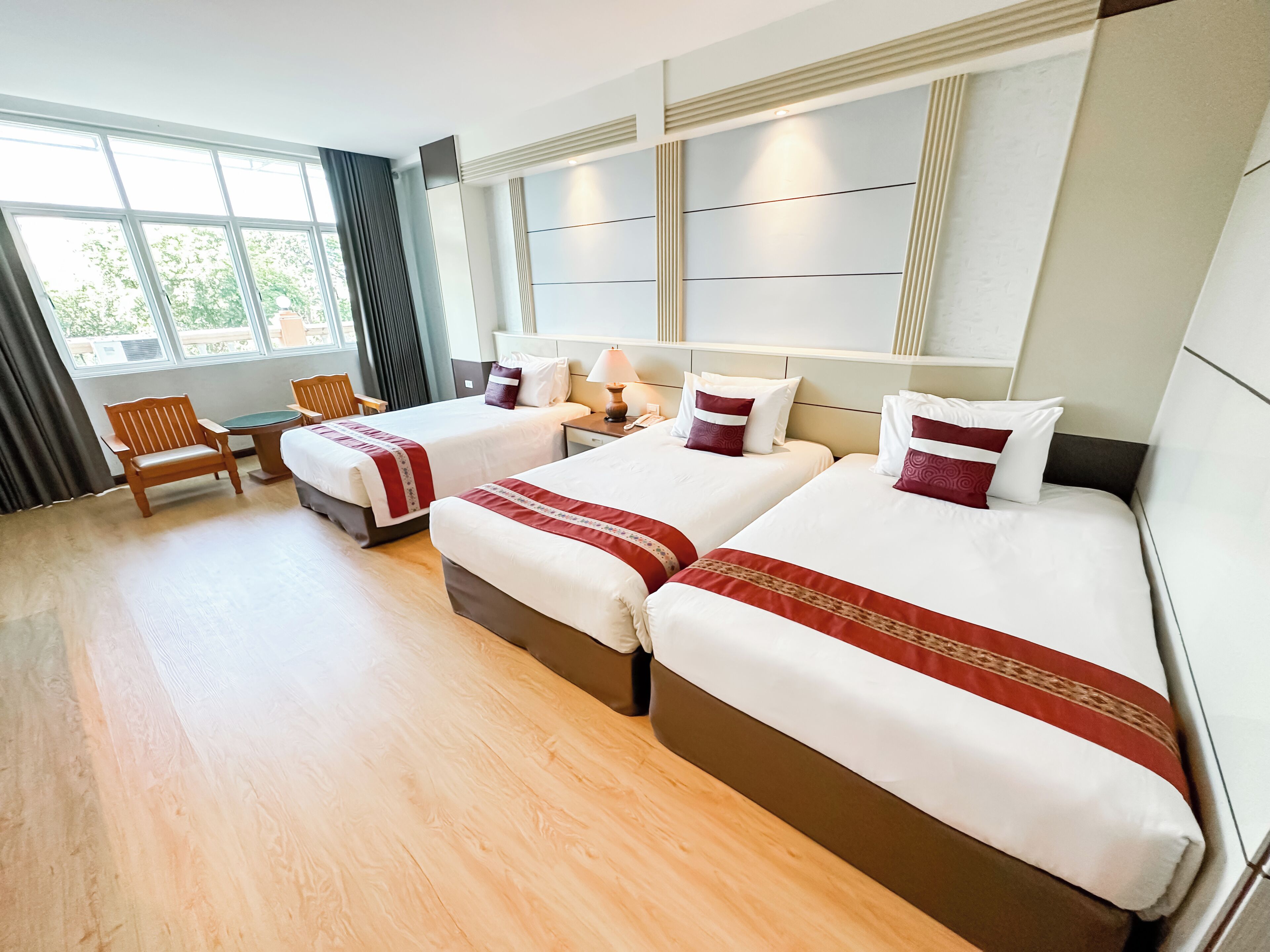 Quarto de hotel com duas camas, uma cadeira de madeira, uma mesinha e uma janela com cortinas.
