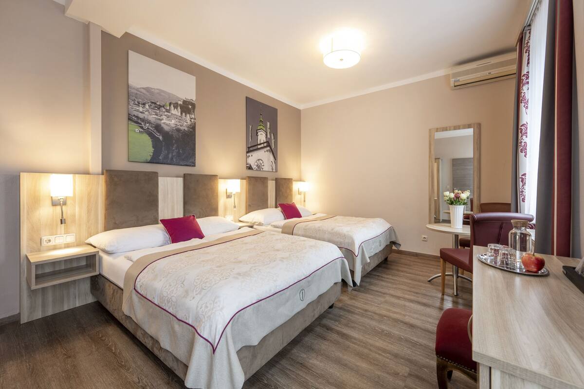 Quarto de hotel com duas camas, uma mesa com uma bandeja de frutas, um espelho e obras de arte nas paredes.