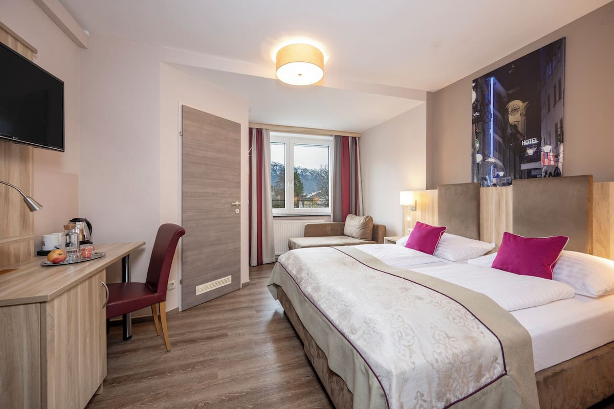 Quarto de hotel com cama, escrivaninha, cadeira, televisão e janela com cortinas.