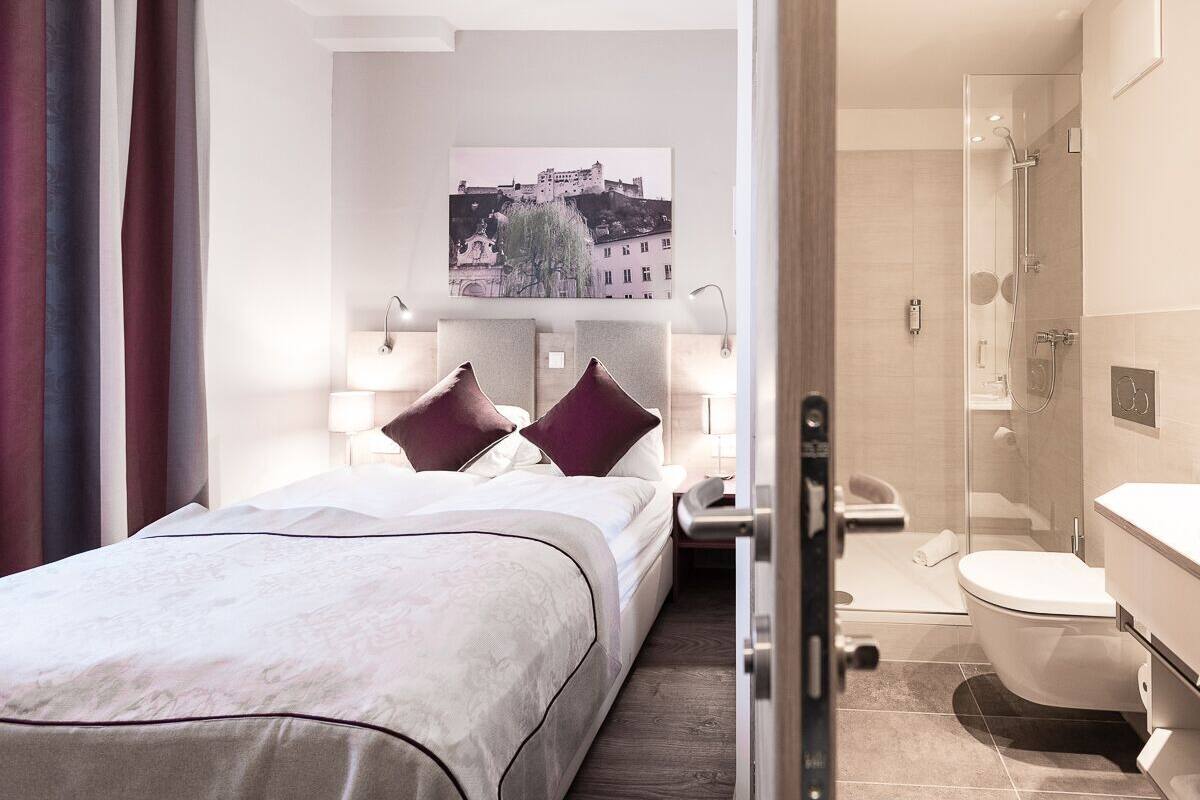 Quarto de hotel com cama, abajures, pintura de um castelo, chuveiro e vaso sanitário.