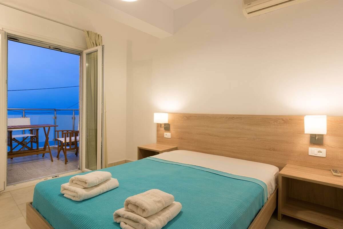 Quarto de hotel com uma cama, duas mesas de cabeceira, uma janela com cortinas e vista para a varanda externa.