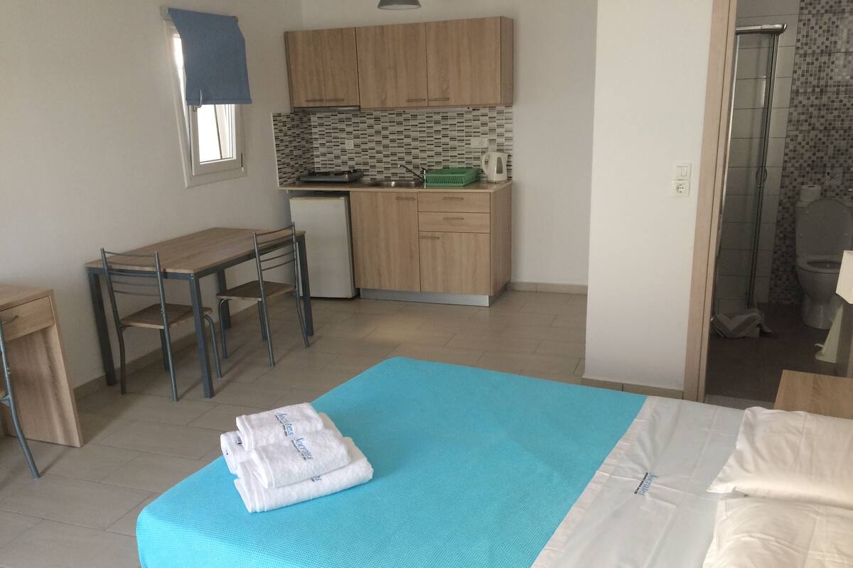 Quarto com cama, escrivaninha, cadeira, pequeno espaço de cozinha e banheiro.