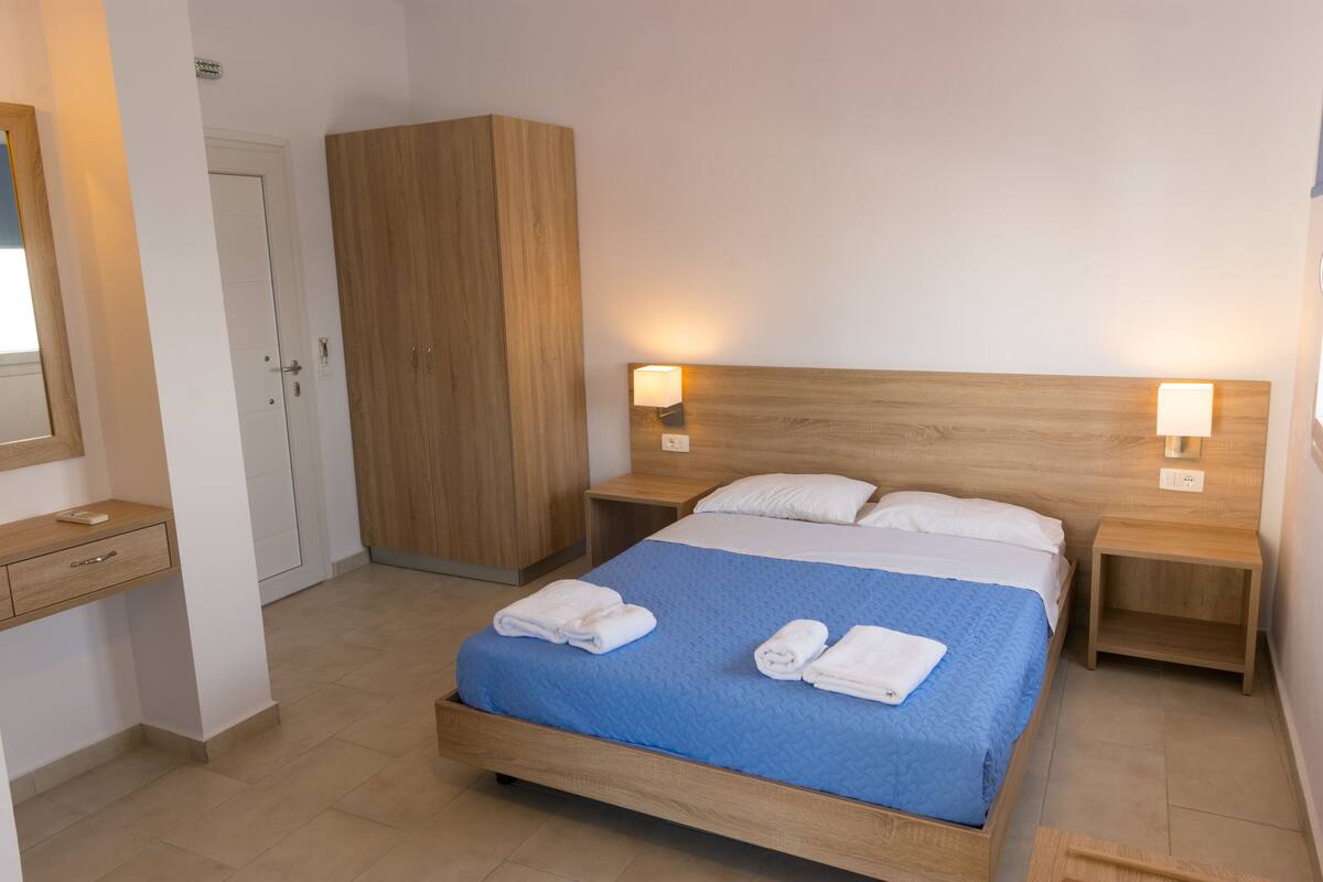 Cama bem arrumada com roupa de cama azul, duas luminárias de cabeceira, um guarda-roupa de madeira e uma pequena mesa de cabeceira.