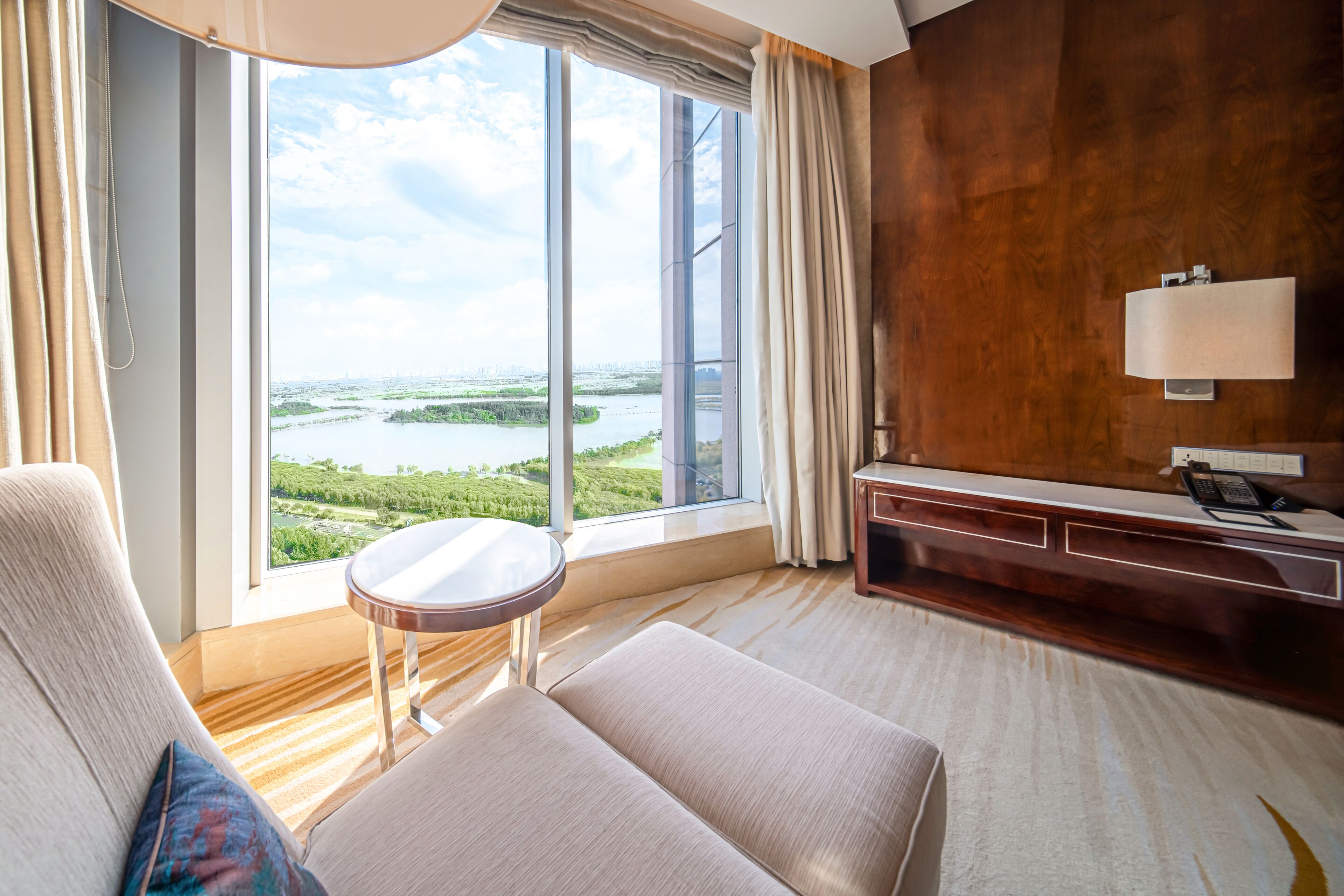 Quarto de hotel com uma janela grande, uma cama, uma mesa pequena e uma escrivaninha com abajur.