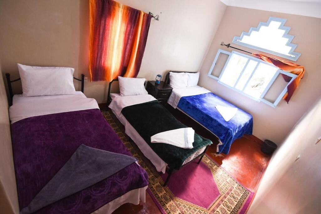 Quarto com três camas, uma janela com cortinas, luminária fixada na parede e um tapete pequeno.