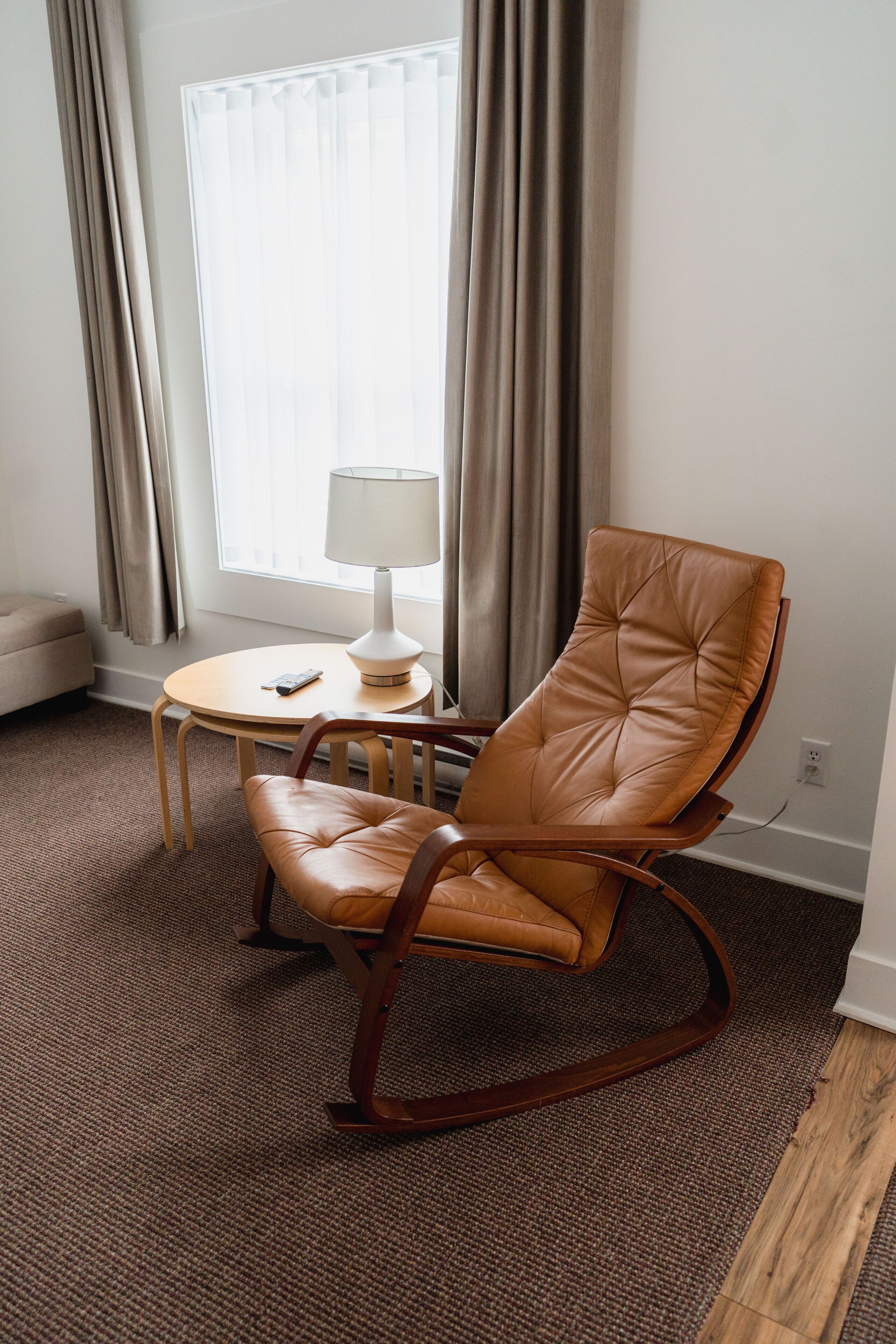 Quarto com poltrona de couro marrom, mesa redonda pequena com abajur e janela com cortinas.