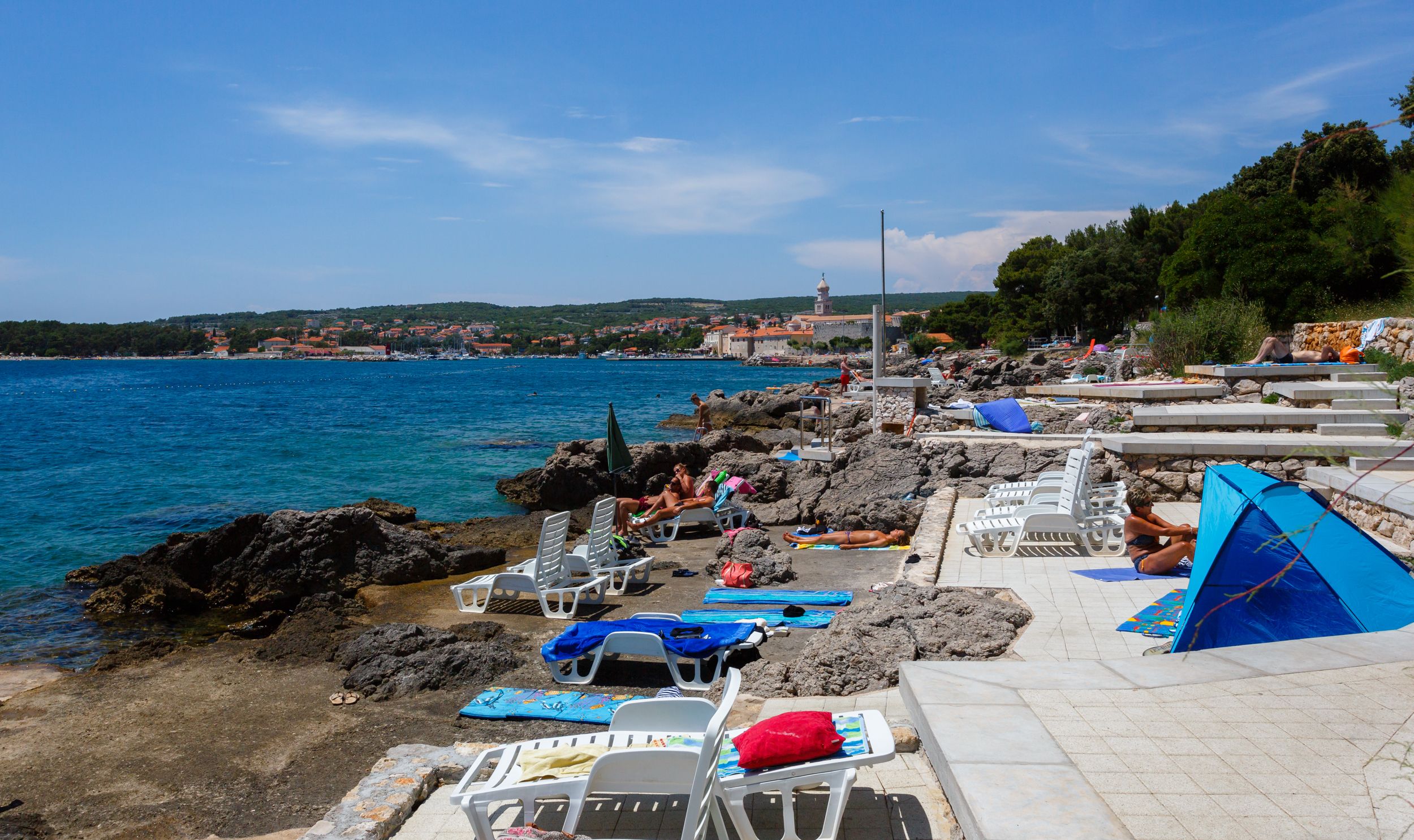 Uma área costeira com costas rochosas, mar azul límpido e vista distante de uma cidade com edifícios de telhado vermelho.