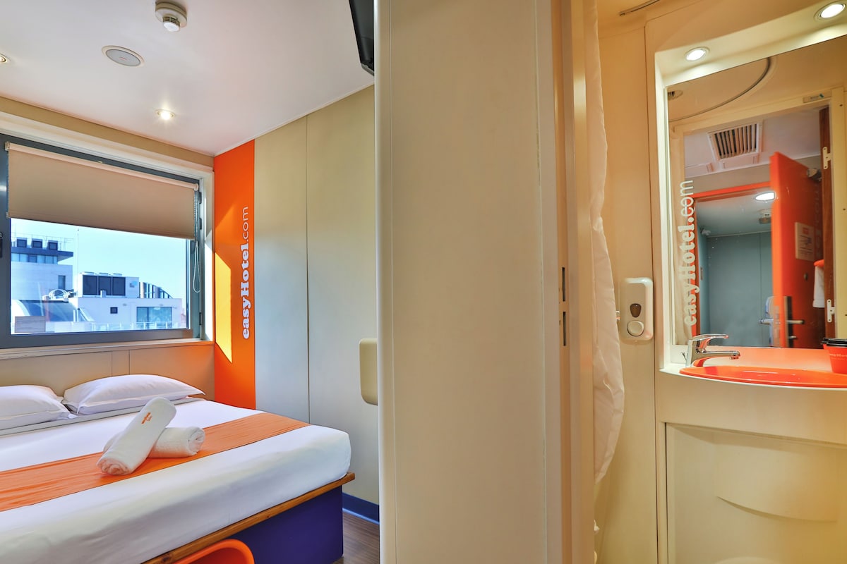 Quarto de hotel com uma cama, janela com vista para edifícios, banheiro com pia e espelho, e porta com maçaneta.