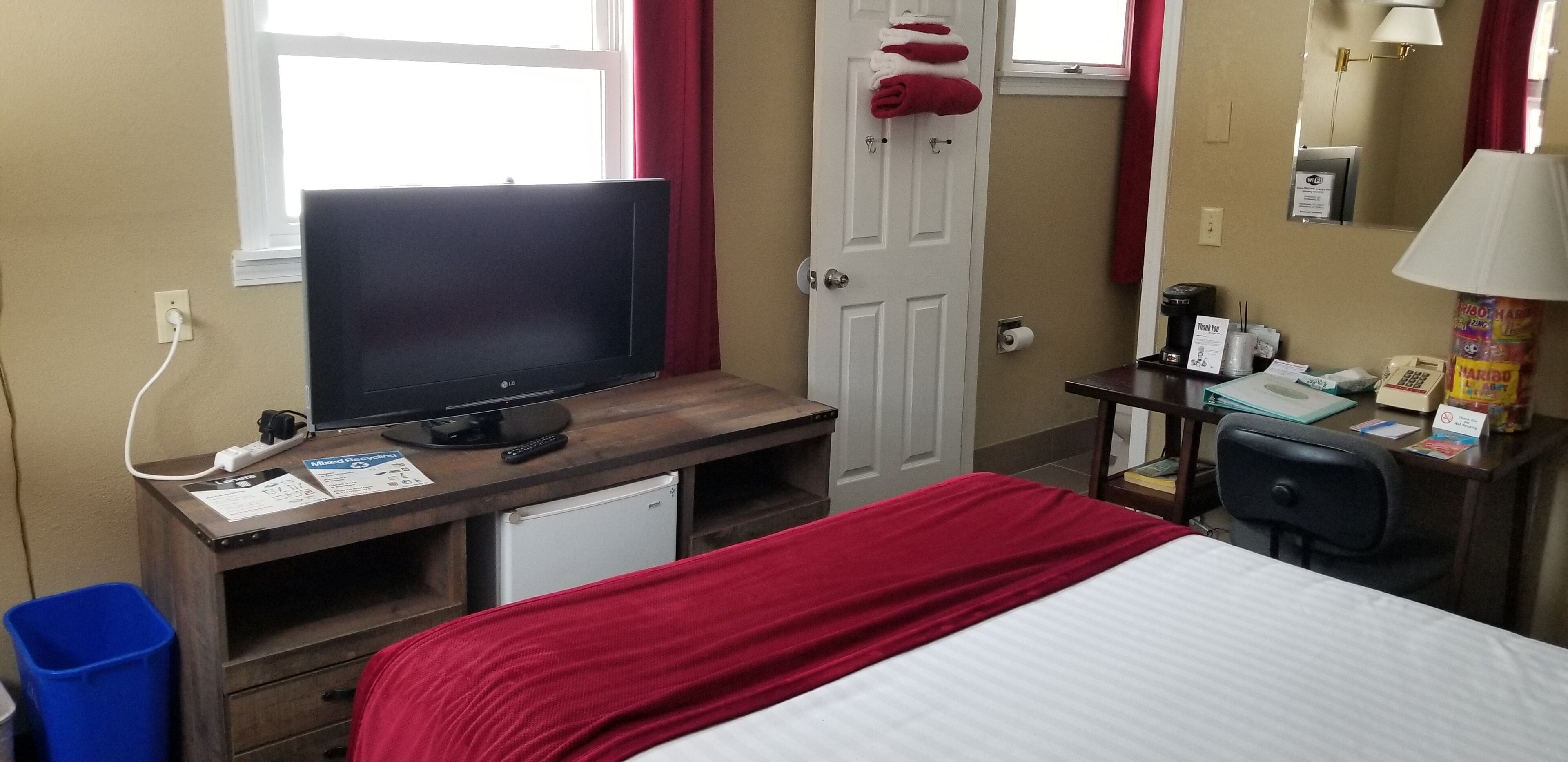 Quarto de hotel com cama, televisão em suporte de madeira, escrivaninha com luminária e pequeno frigorífico.