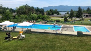 Área da piscina com vista para montanhas e um lago, rodeada por árvores e um campo gramado.
