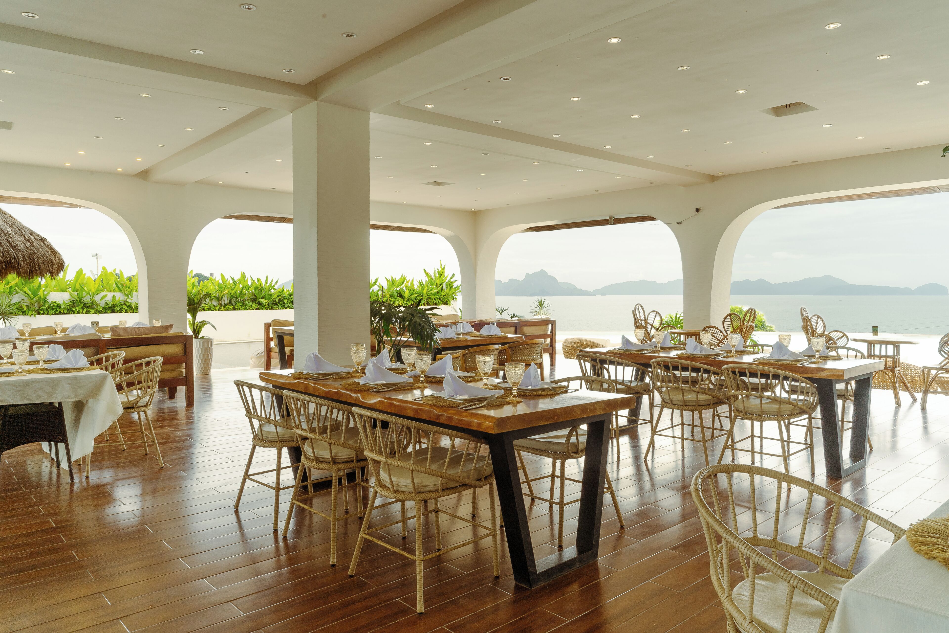 Restaurante com mesas de madeira e cadeiras de vime, amplas janelas com vista para o mar e um telhado de palha.