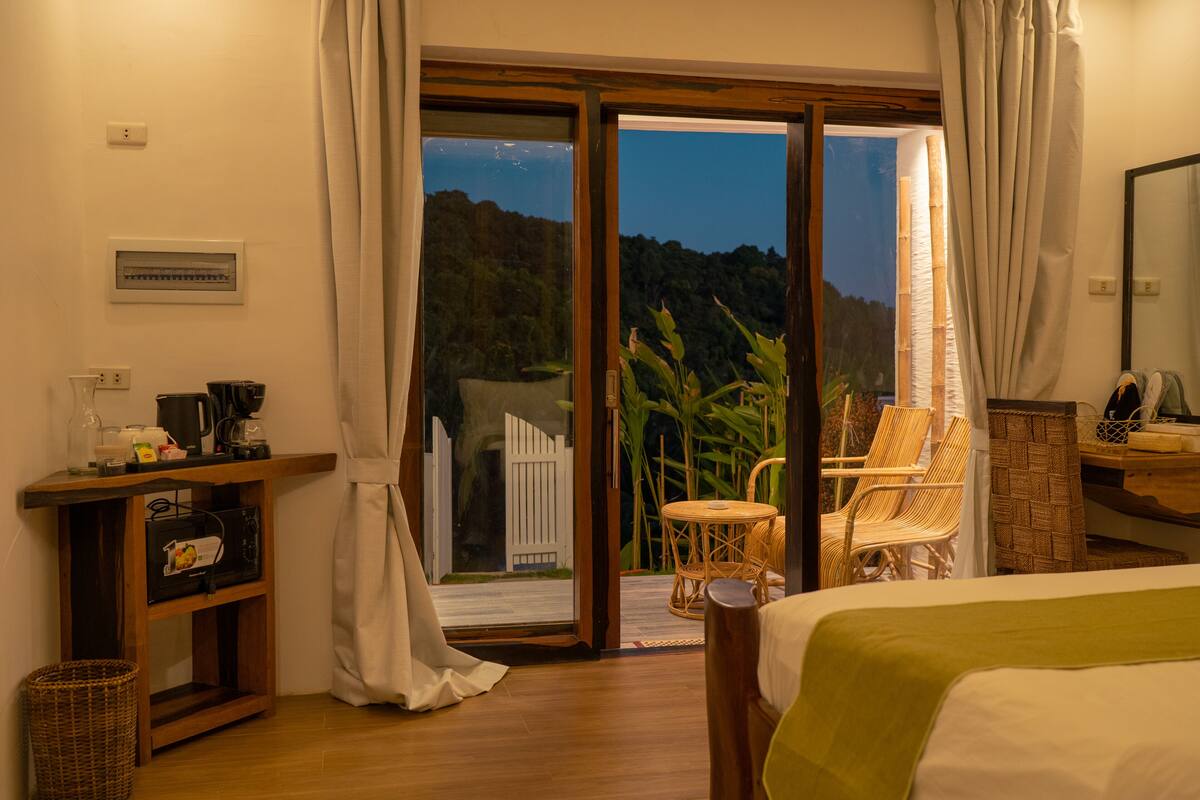 Quarto de hotel com cama, uma pequena mesa com cafeteira, uma cadeira de vime e vista para um jardim exuberante através de uma porta de vidro deslizante.