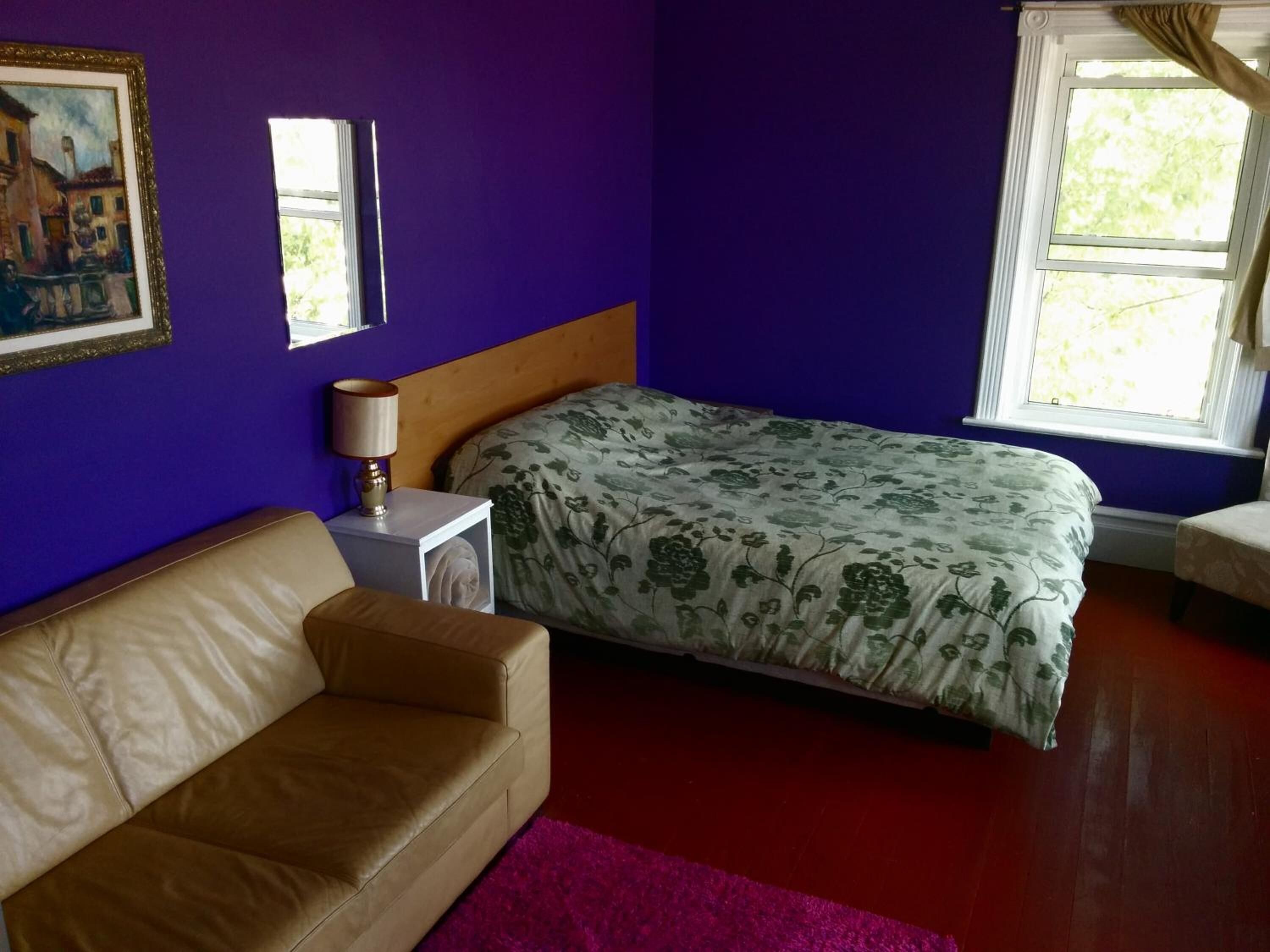 Quarto com cama, mesa de cabeceira, luminária, janela com cortinas, parede roxa e sofá de couro marrom.