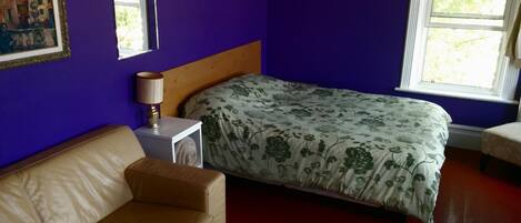 Quarto com cama, mesa de cabeceira, luminária, janela com cortinas, parede roxa e sofá de couro marrom.