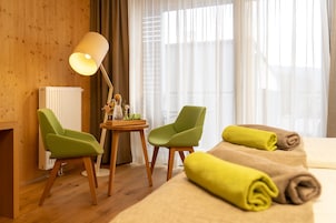 Quarto de hotel com paredes de madeira, uma cama com toalhas dobradas, duas cadeiras verdes, uma pequena mesa com abajur e uma janela com cortinas translúcidas.
