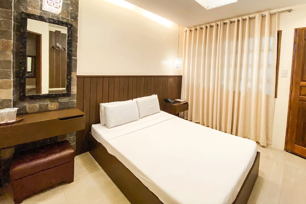 Quarto de hotel com cama, cabeceira de madeira, mesa de cabeceira, espelho, porta de madeira e janela com cortinas.