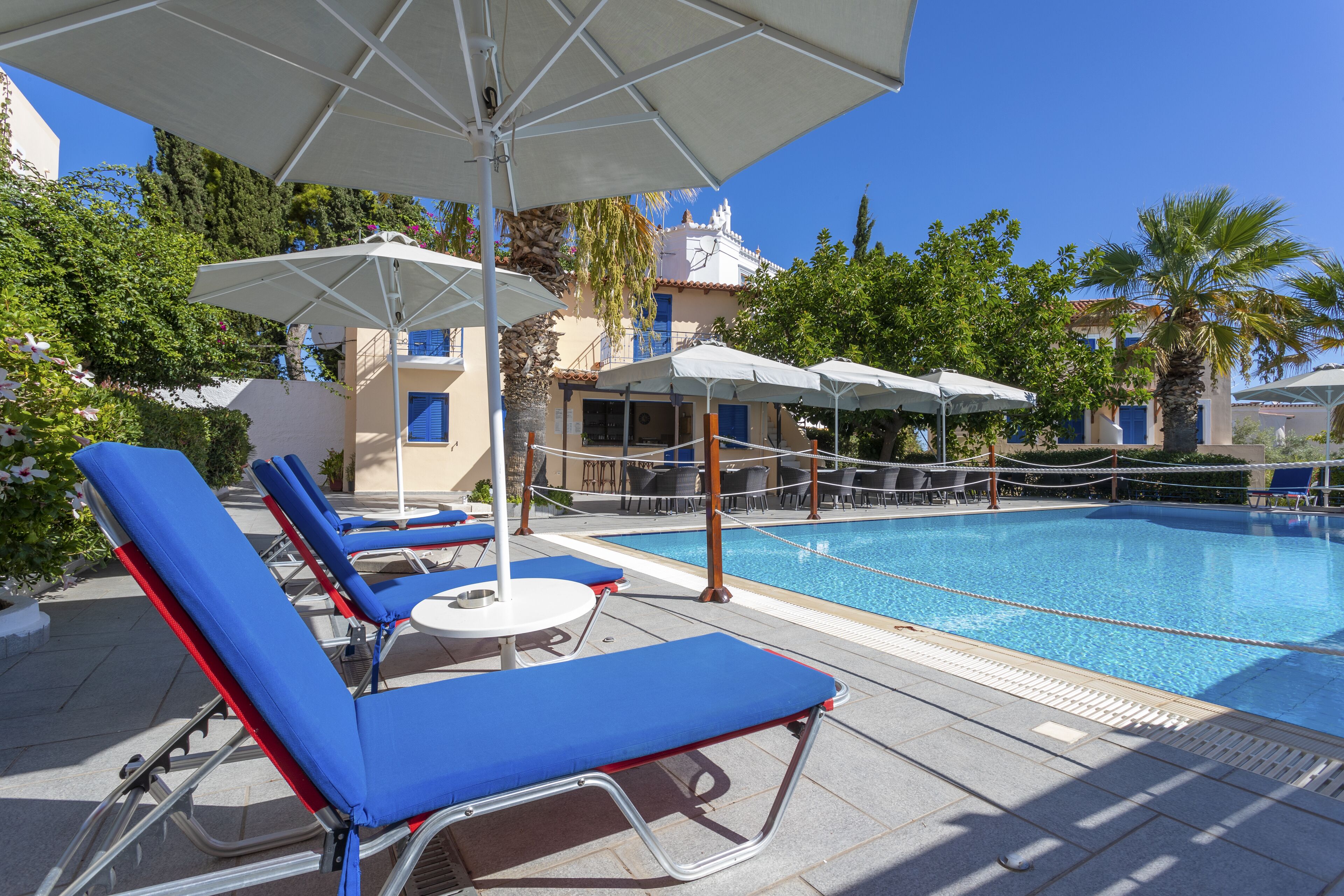 Uma área à beira da piscina com espreguiçadeiras e guarda-sóis, uma piscina azul cristalina e um edifício com uma porta azul ao fundo.