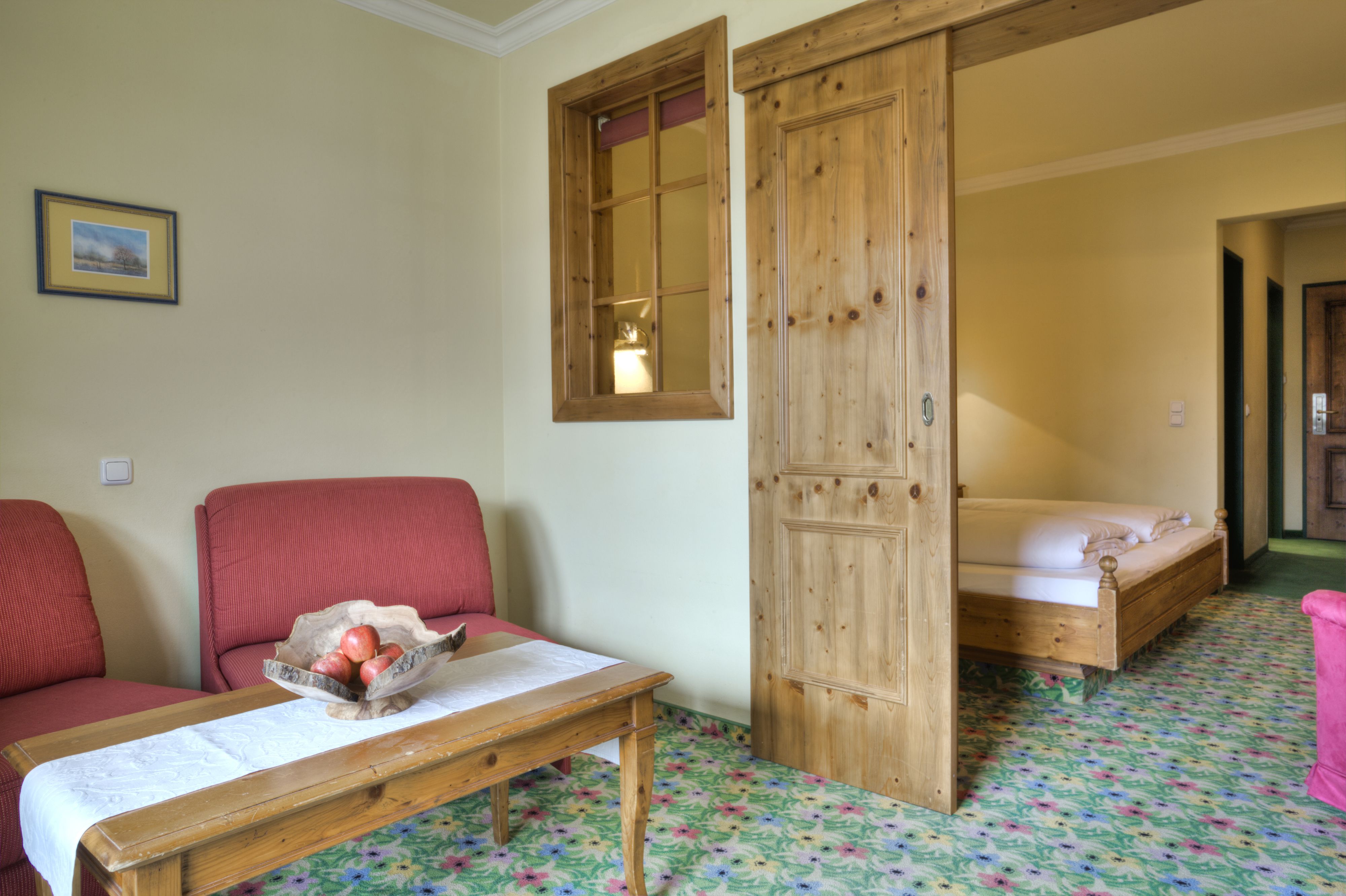 Quarto com cama de madeira, uma cadeira vermelha, uma mesa de madeira com toalha branca e uma janela com moldura de madeira.