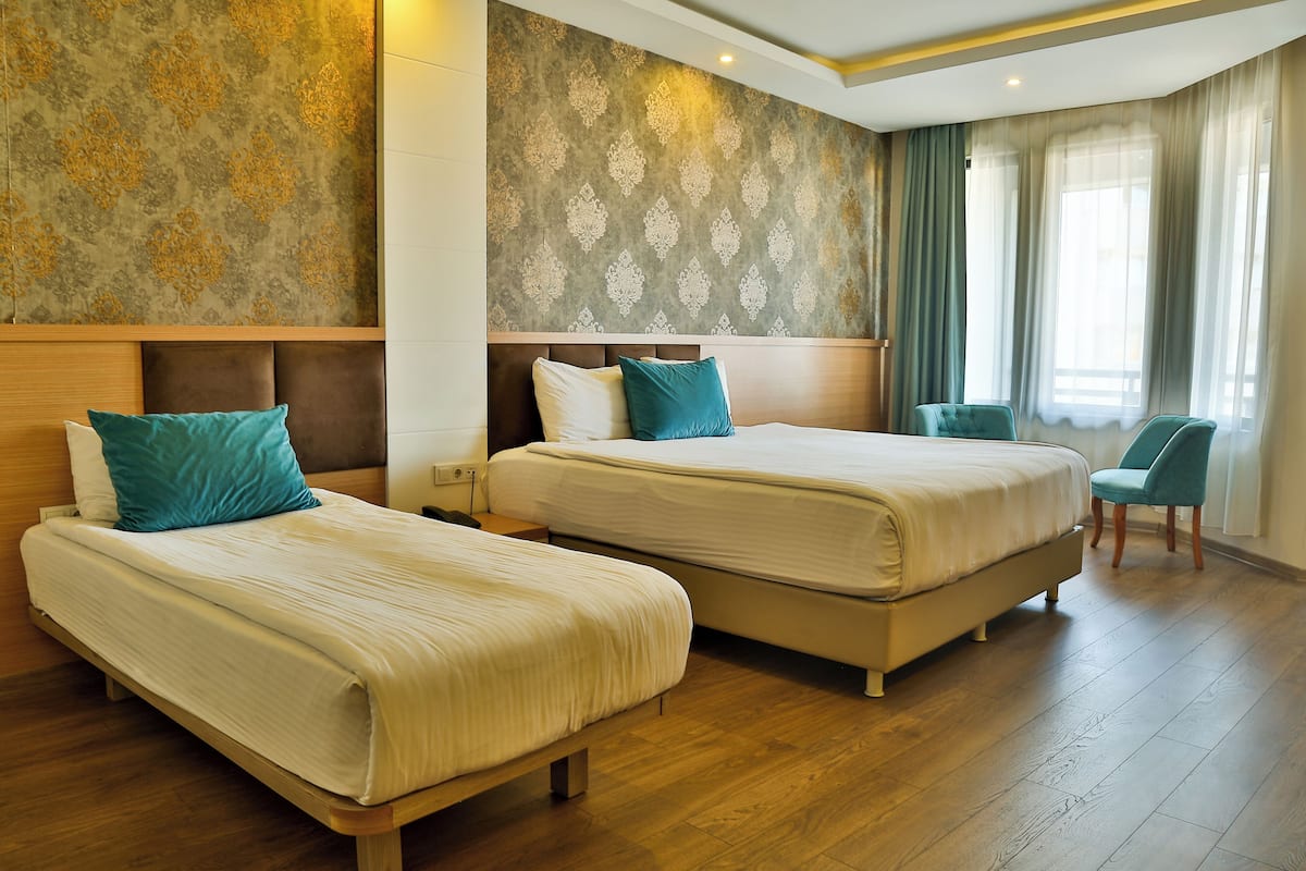 Quarto de hotel com duas camas, cabeceira de madeira, uma cadeira verde-água e uma janela grande com cortinas.